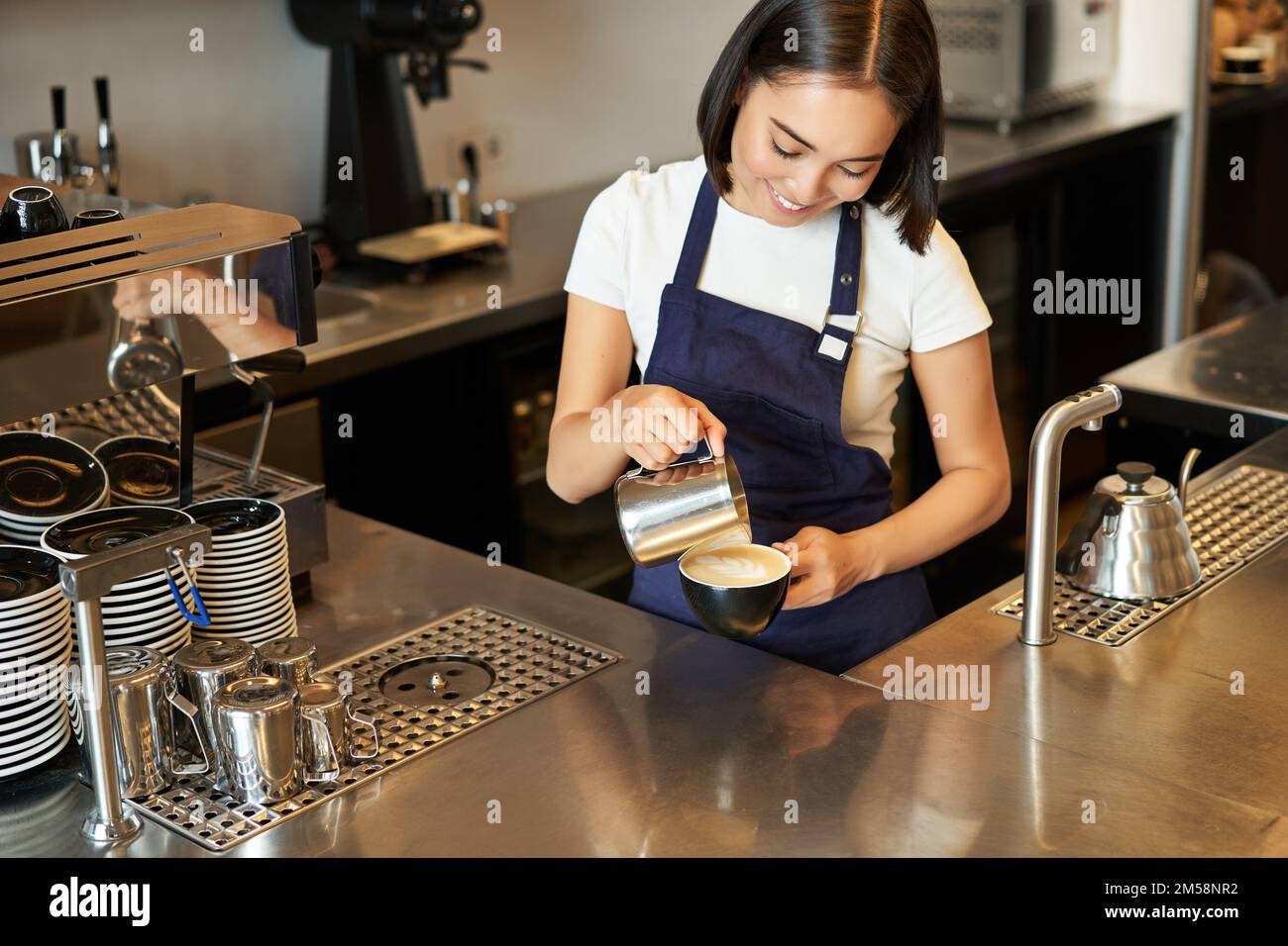 Smiling asian girl barista, cafe staff pouring steamed milk in coffee ...