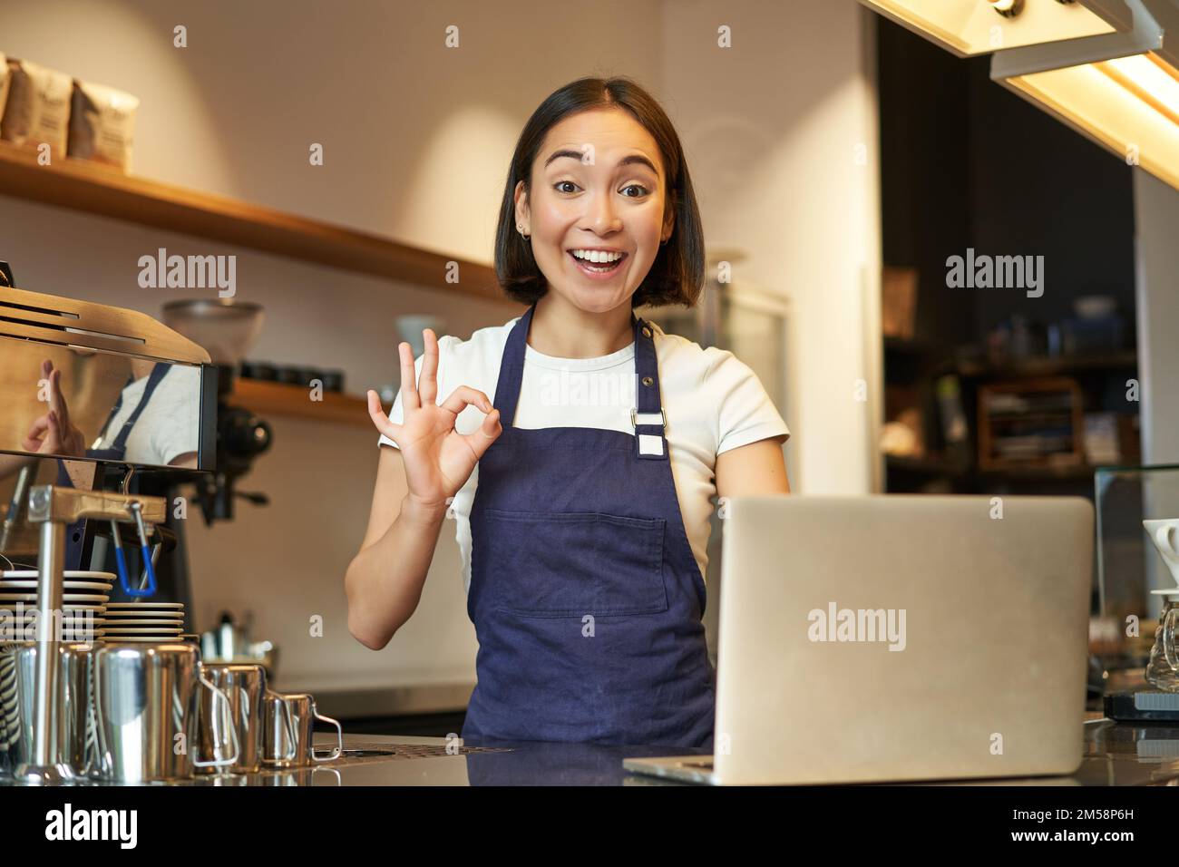 Portrait of barista girl, asian female cafe worker, shows okay, ok sign ...