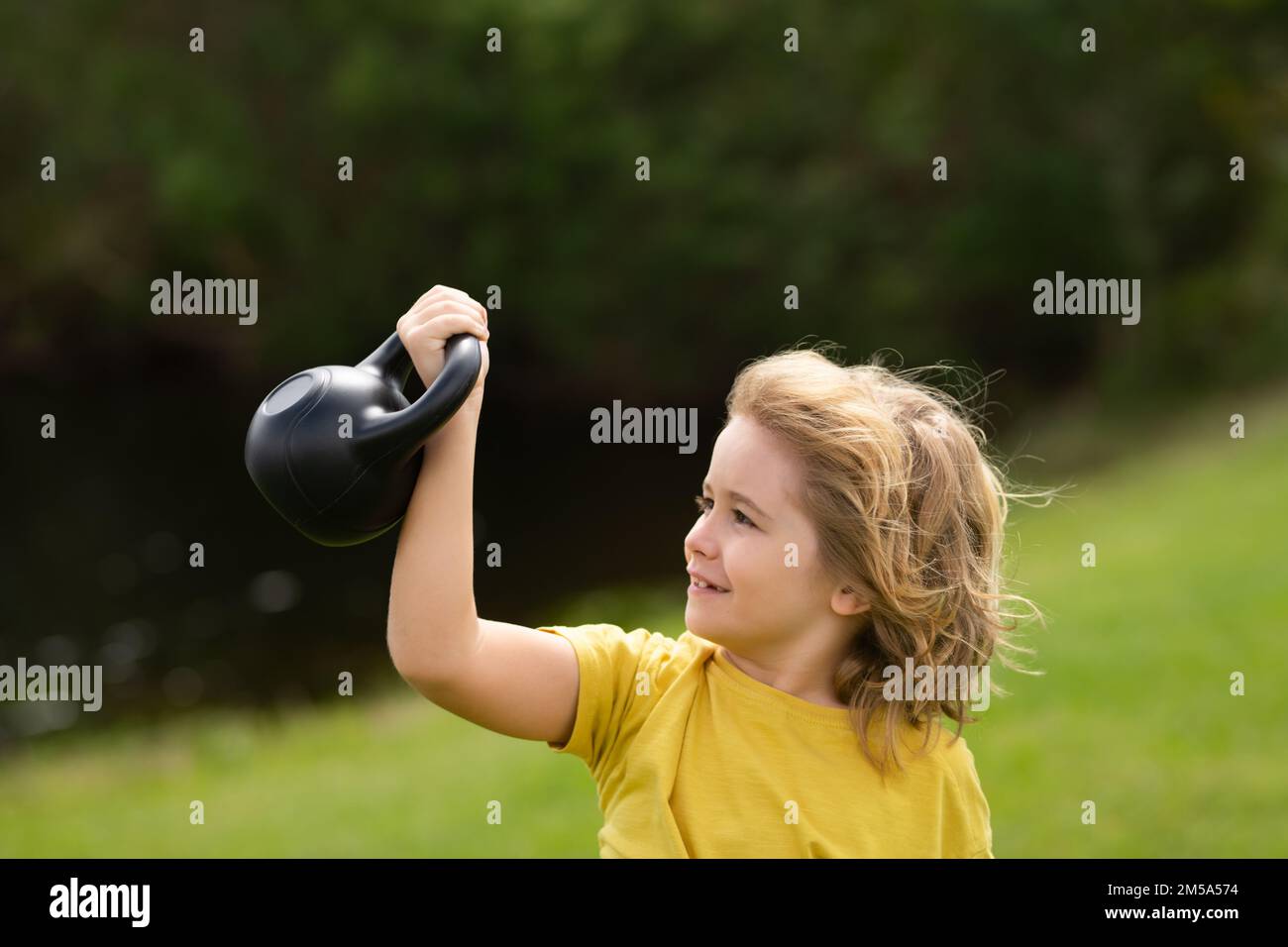 Kid lifting the kettlebell in park outside. Child boy pumping up biceps ...