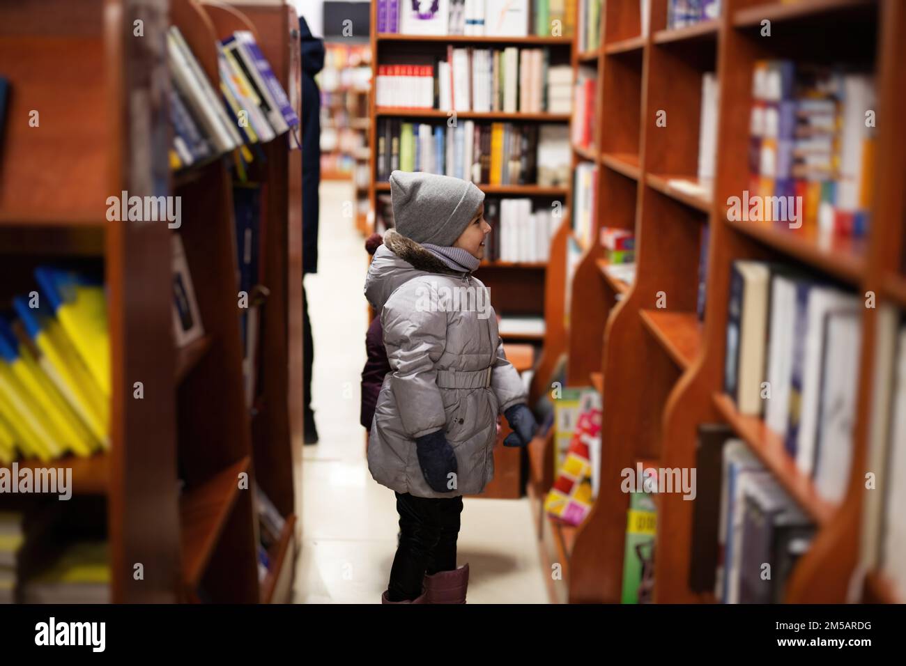 Little cute girl in jacket reaching a book from bookshelf at the ...