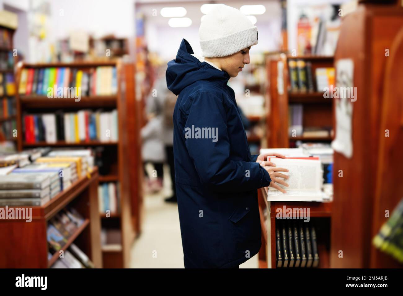 Teenager boy in jacket reaching a book from bookshelf at the library ...