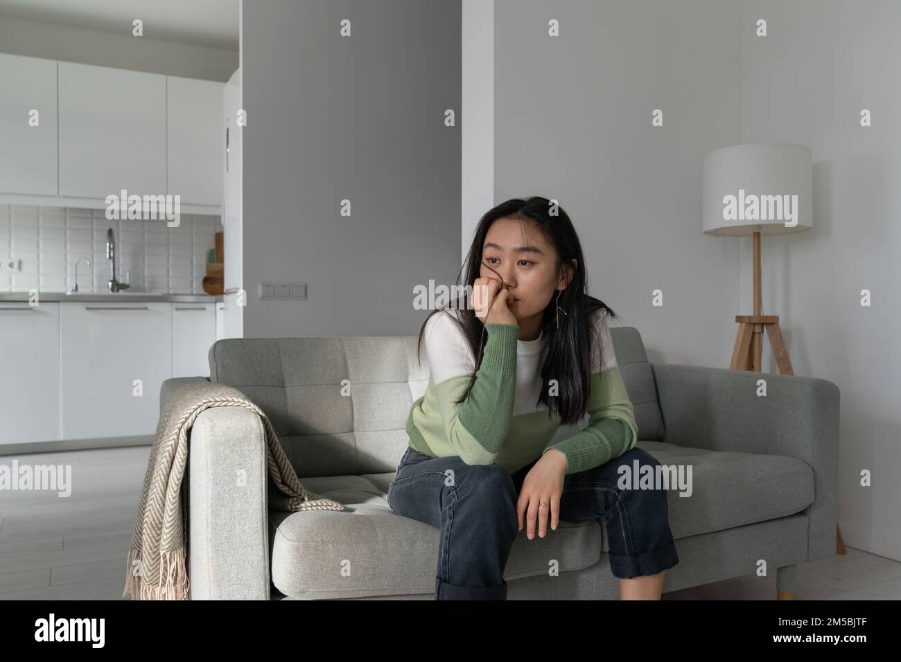 Upset unhappy Asian woman alone sits on couch propping head with hand ...