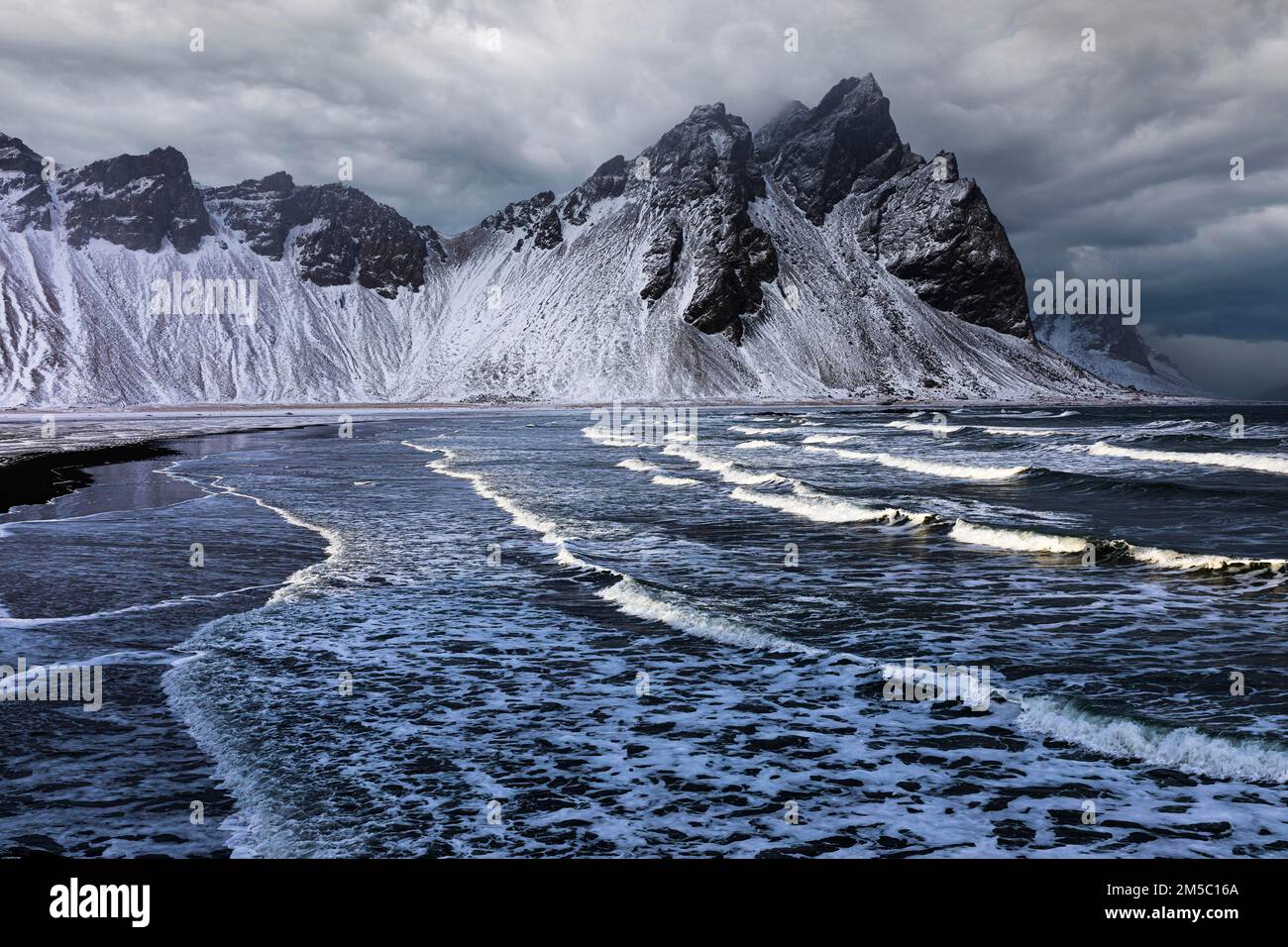 White foamy waves on the beach of Stokkness, behind snowy rocky slopes ...