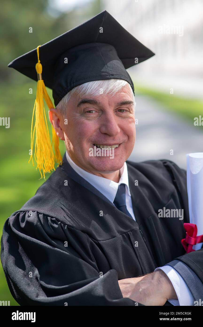 Portrait of an elderly man in a graduation gown and with a diploma in ...