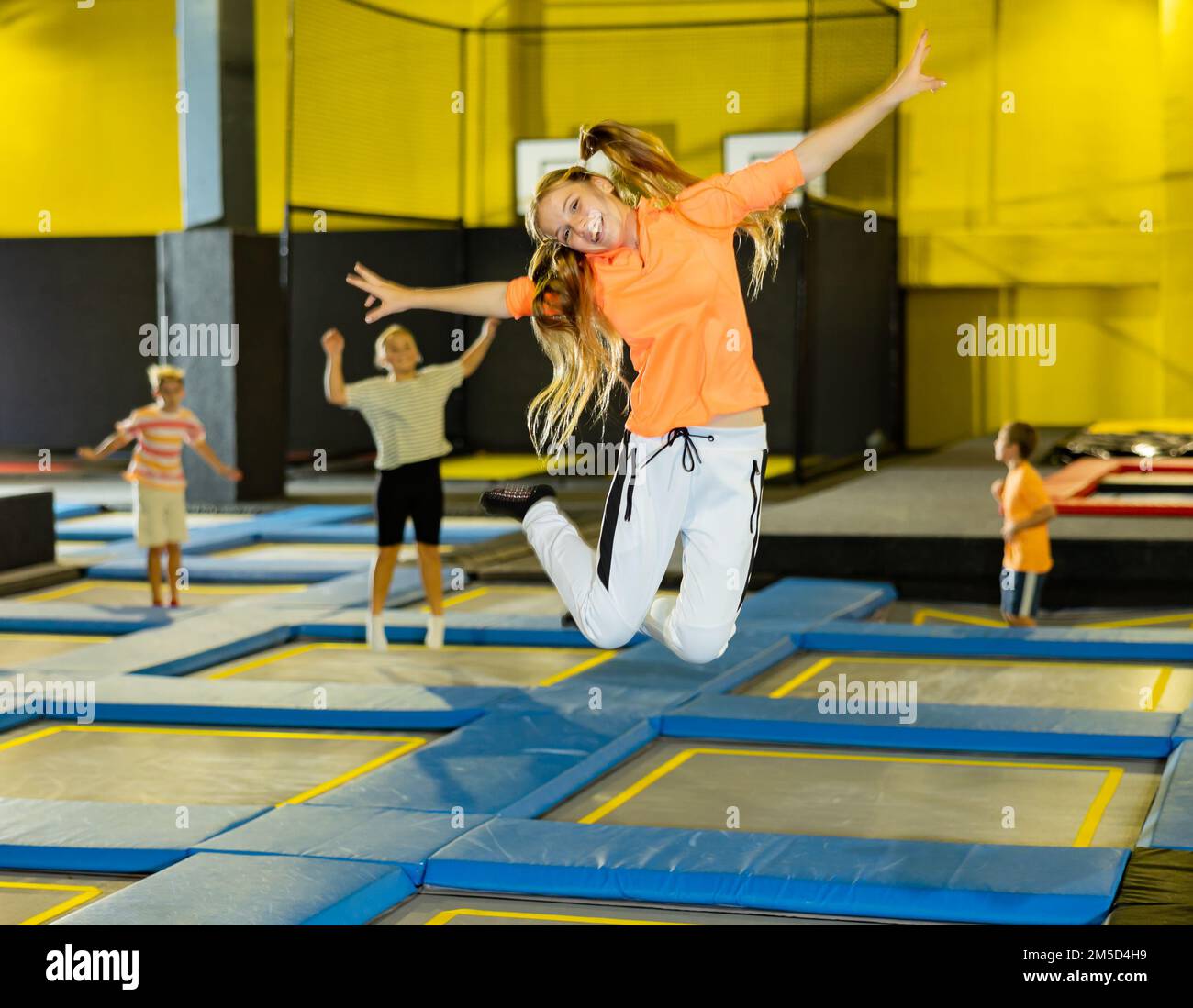 Excited teen girl in orange sports jumper and white pants jumping and indulge on trampolines in ...