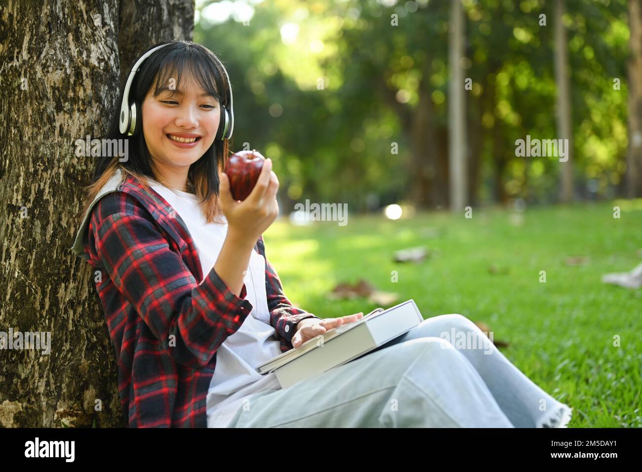 Pretty young asian woman eating apple under huge tree on green grass at ...