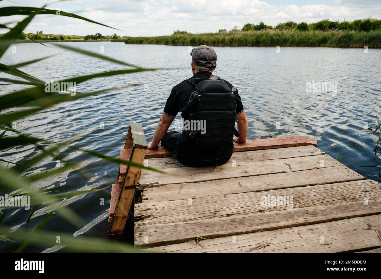 A man sits on the edge of a wooden bridge near a lake next to reeds, a ...