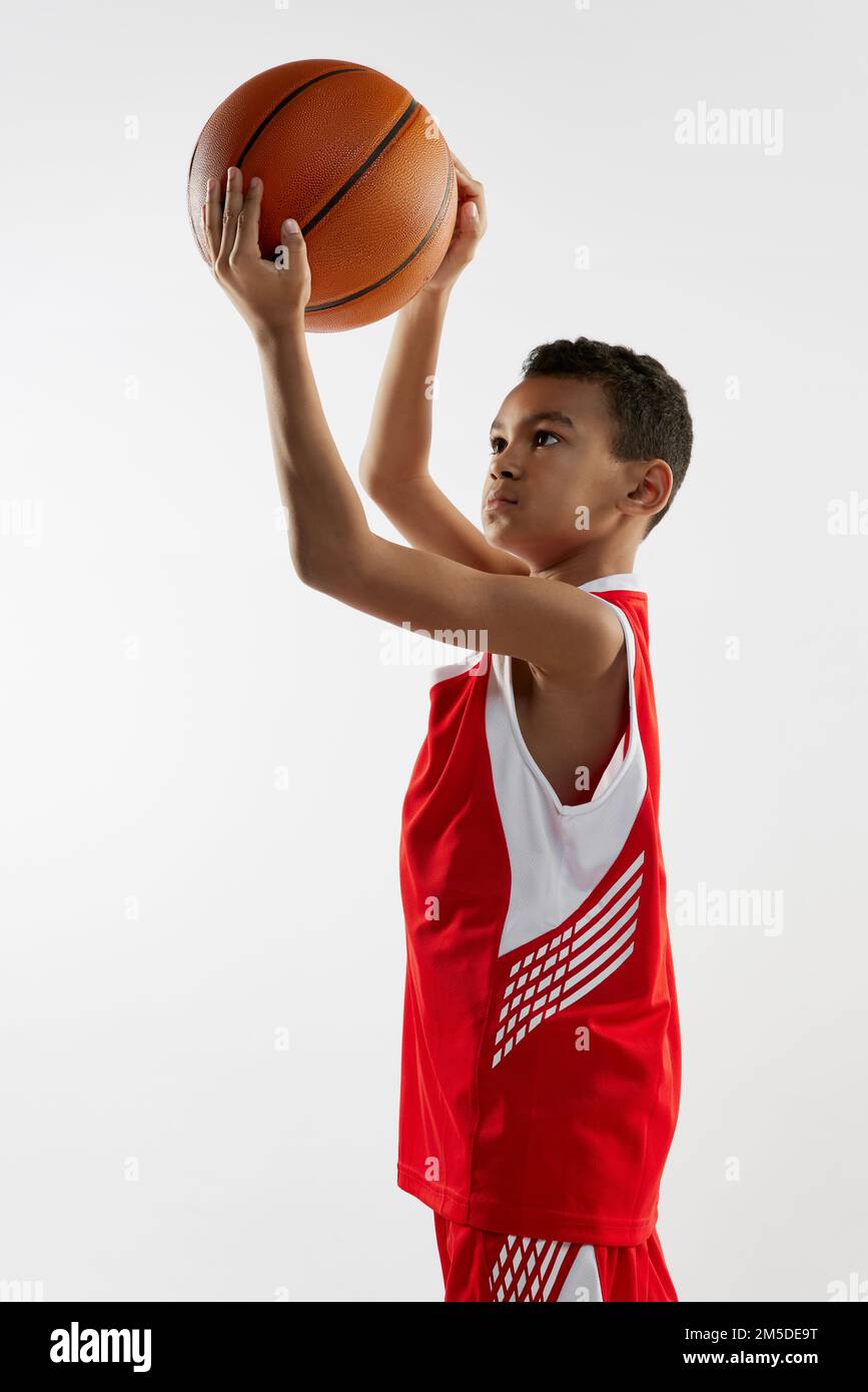 Portrait of boy in red uniform training, playing basketball over grey ...