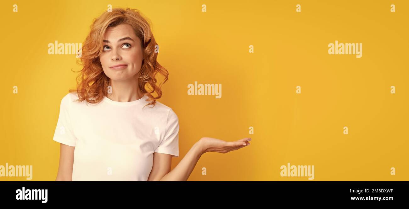 pondering redhead woman presenting product on yellow background with copy space. Beautiful woman ...