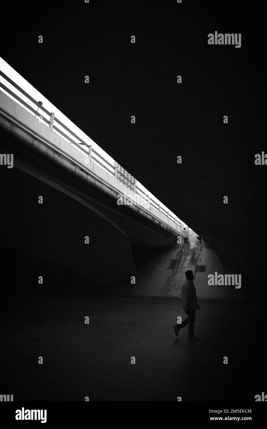 A grayscale of a man walking on a pedestrian walkway under a bridge ...