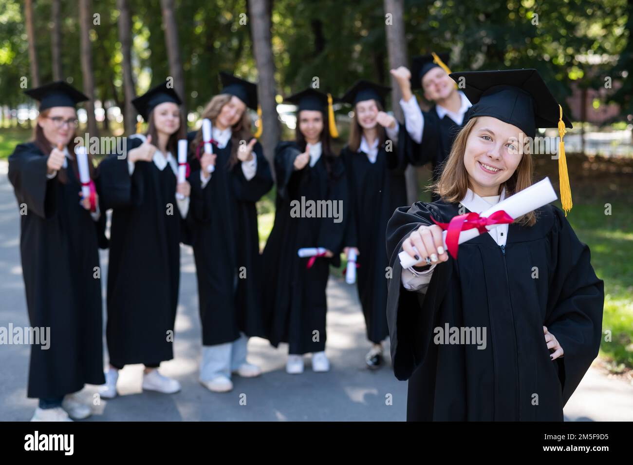 Group of happy students in graduation gowns outdoors. A young girl with ...