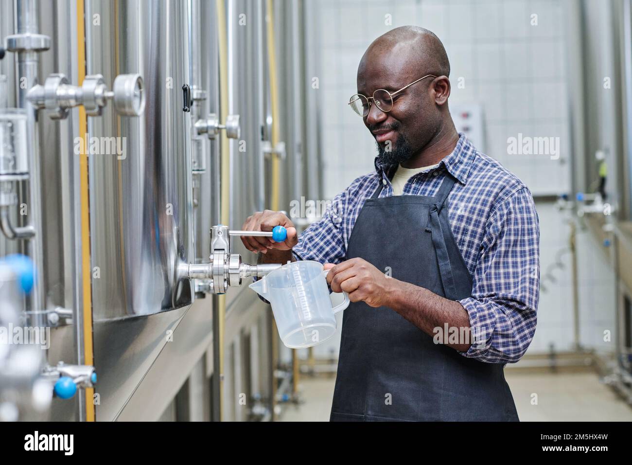African American worker pouring fresh beer to taste it after brewing ...