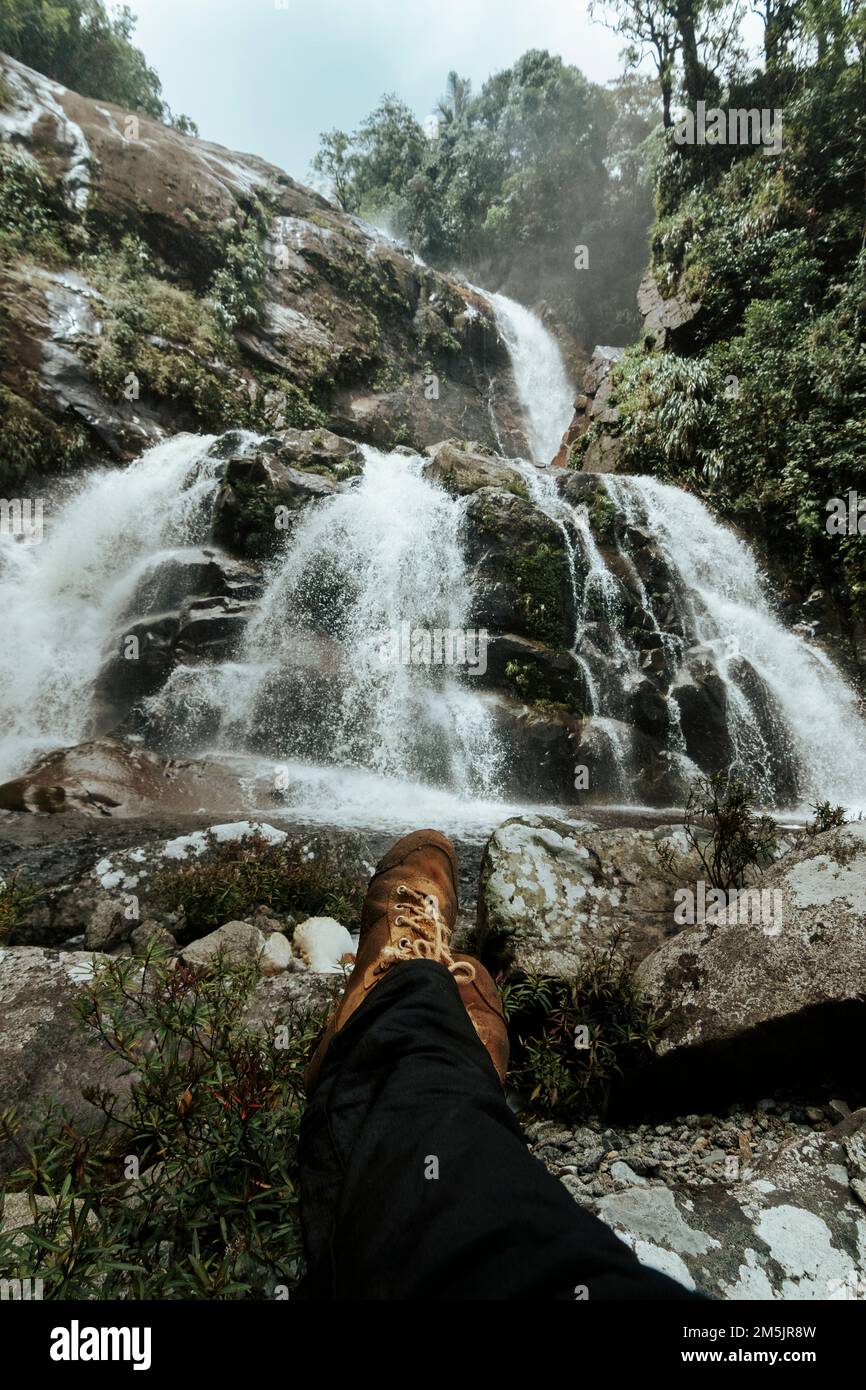 A vertical shot of a feet and shoes of a man with a scenic waterfall on ...