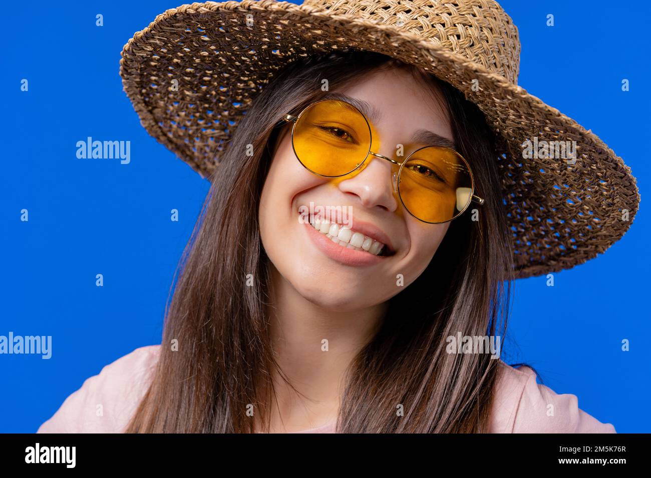 Charming woman in yellow eyewear and straw hat on blue studio backdrop ...