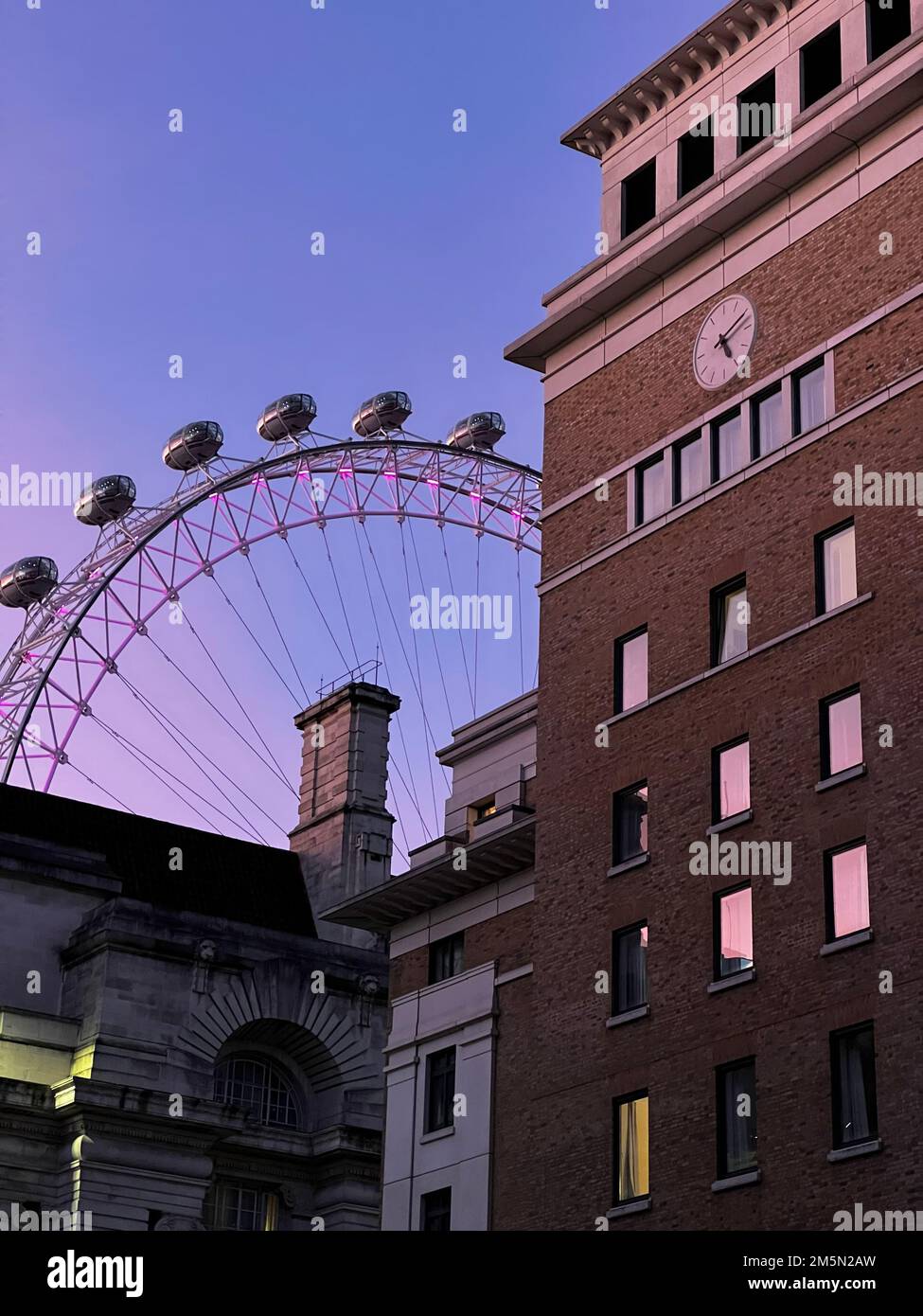 A vertical shot of the London eye behind the city buildings with a blue ...