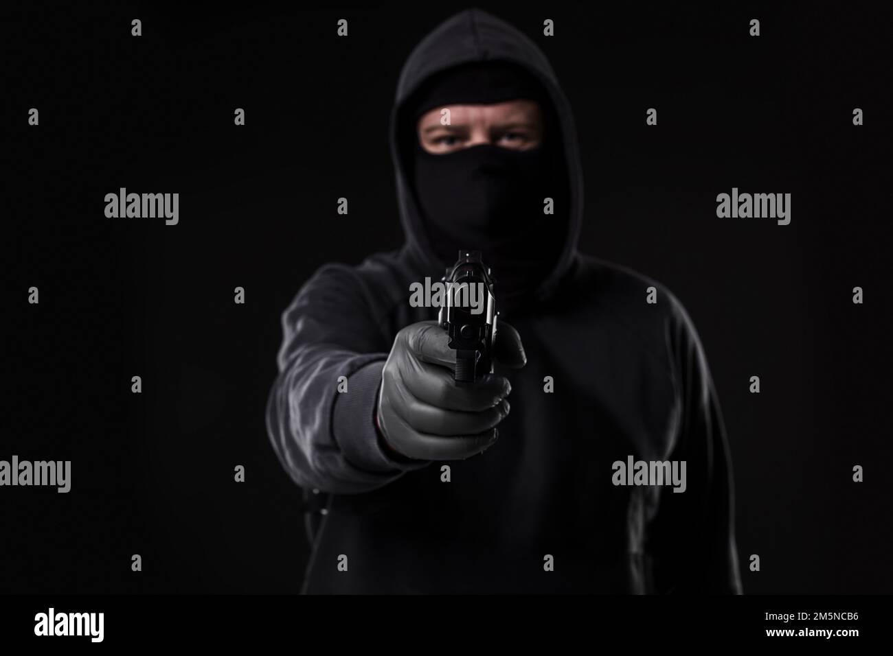 Masked robber with gun aiming into the camera against a black ...