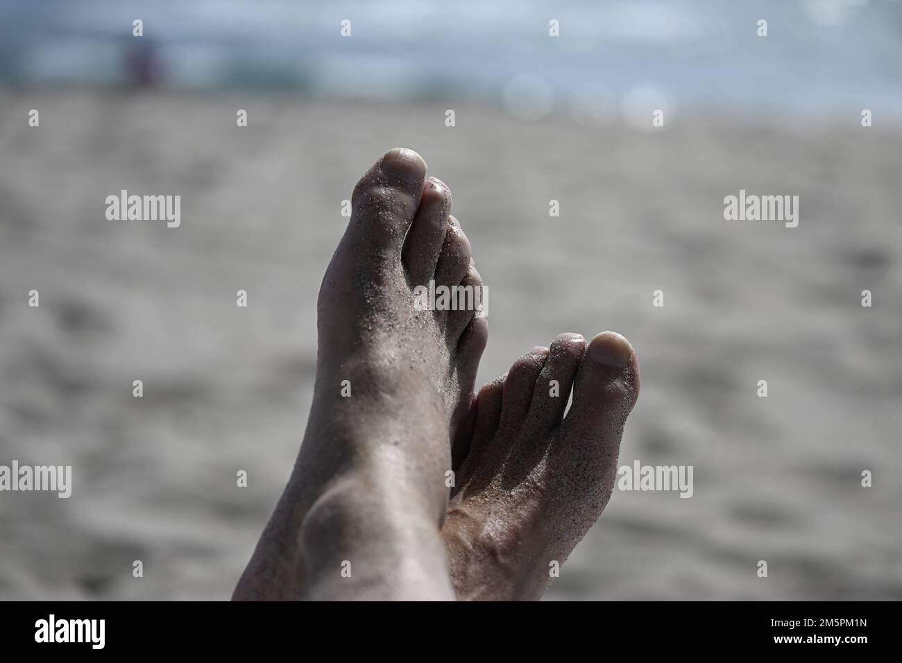 A closeup of a men's toes covered in sand at the beach with a blurred ...