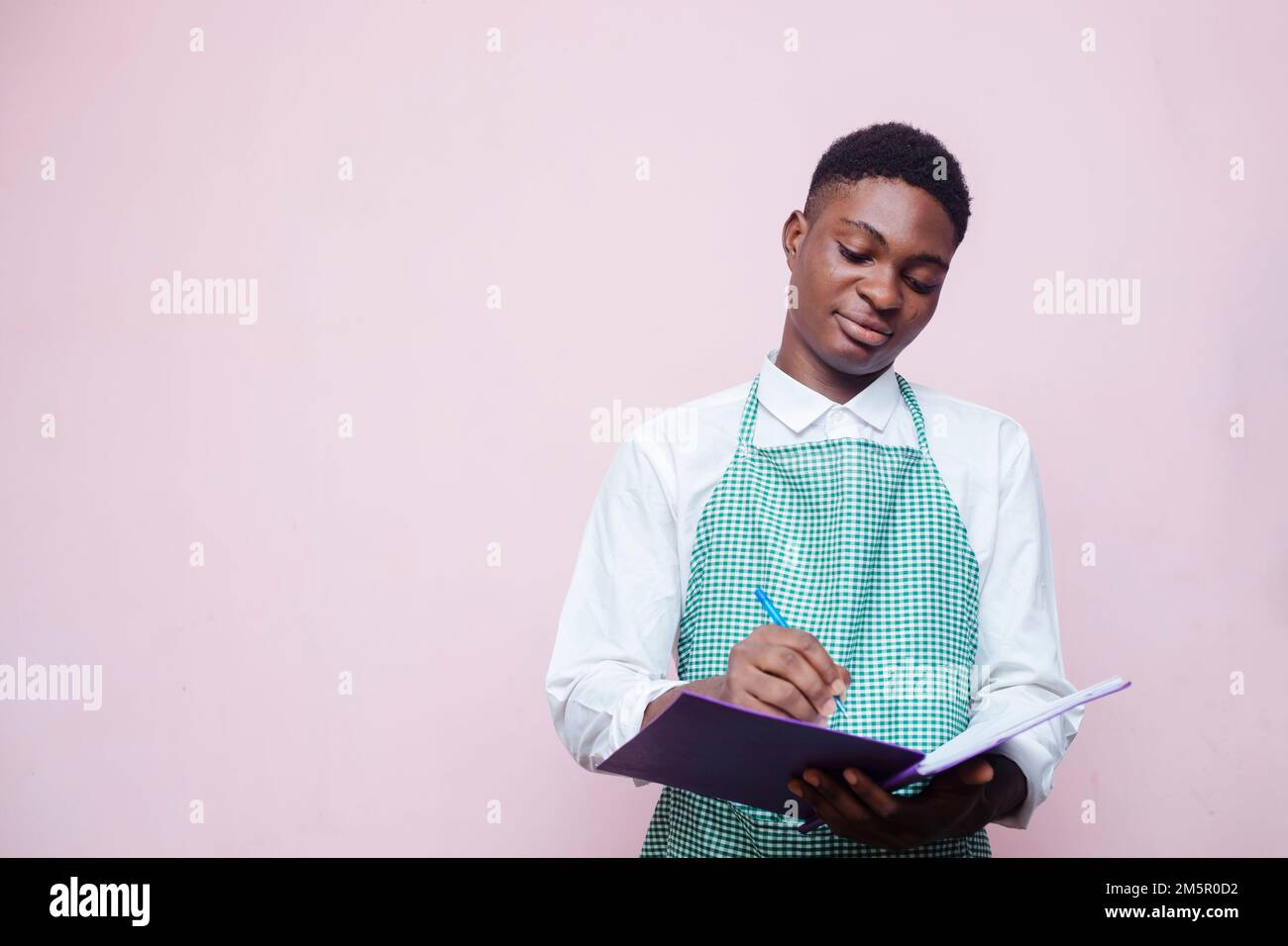 An African man wearing apron and making notes in notebook in white ...