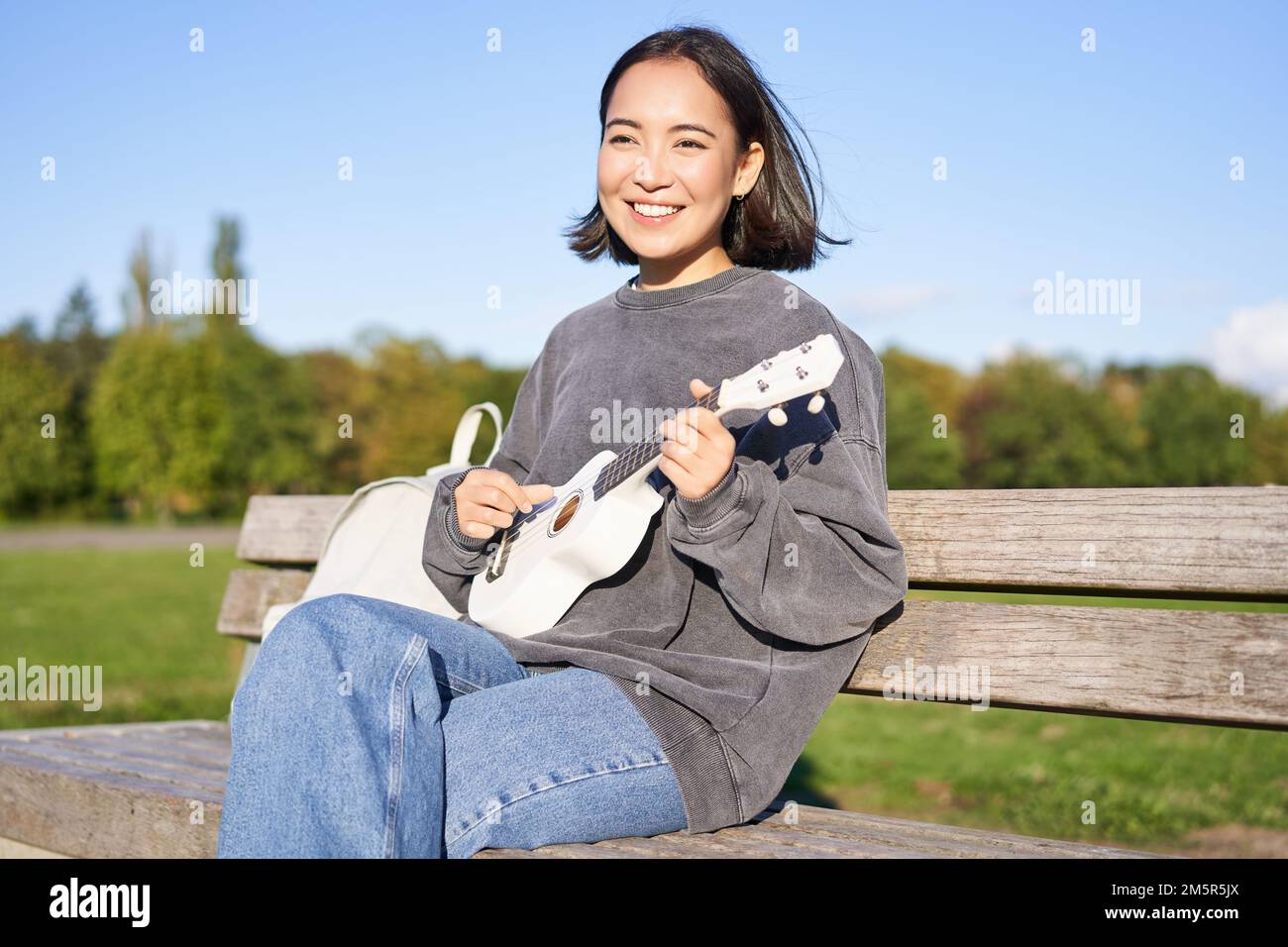 Happy cute girl sits alone on bench in park, plays ukulele guitar and ...