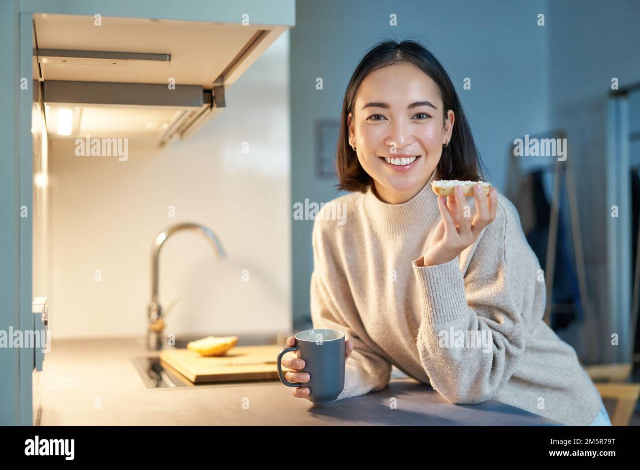 Beautiful young asian woman eating toast on her kitchen, drinking ...