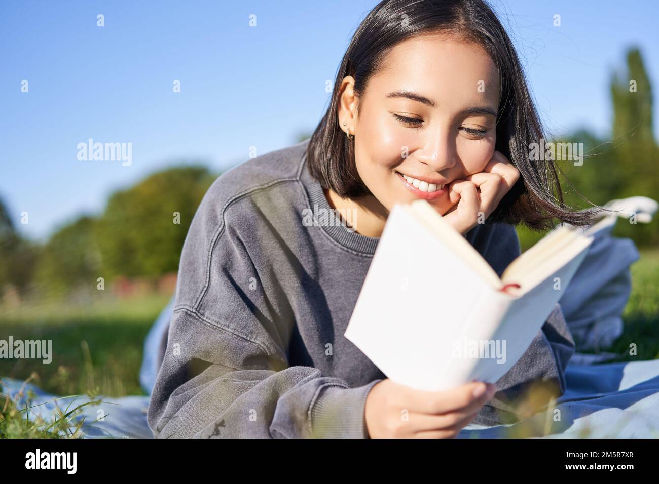 Portrait of beautiful smiling asian girl, reading in park, lying on ...
