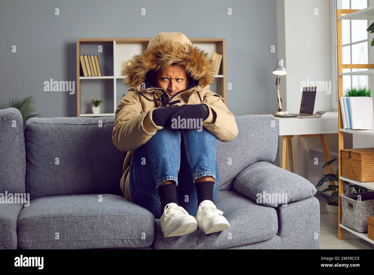 Young frozen indian man sitting in a winter coat on the sofa at home ...