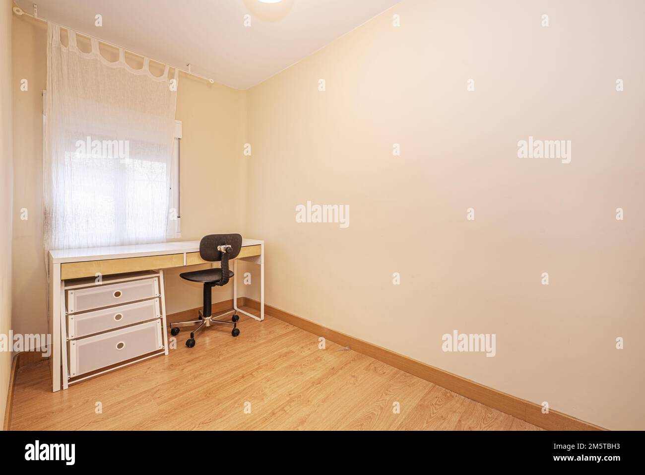 A mostly empty room with a white study table with a chest of drawers ...