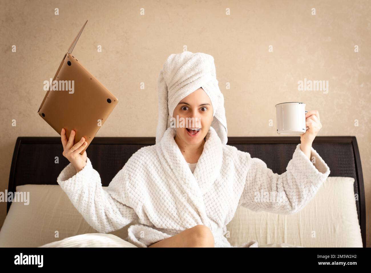 surprised woman in a white dressing gown. in one hand holds a laptop in ...