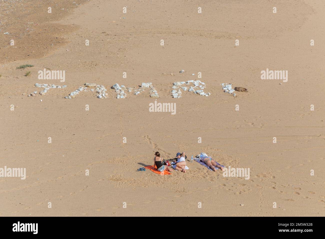 Sunbathers on sandy beach by sign written in rocks, Tamri, Morocco ...
