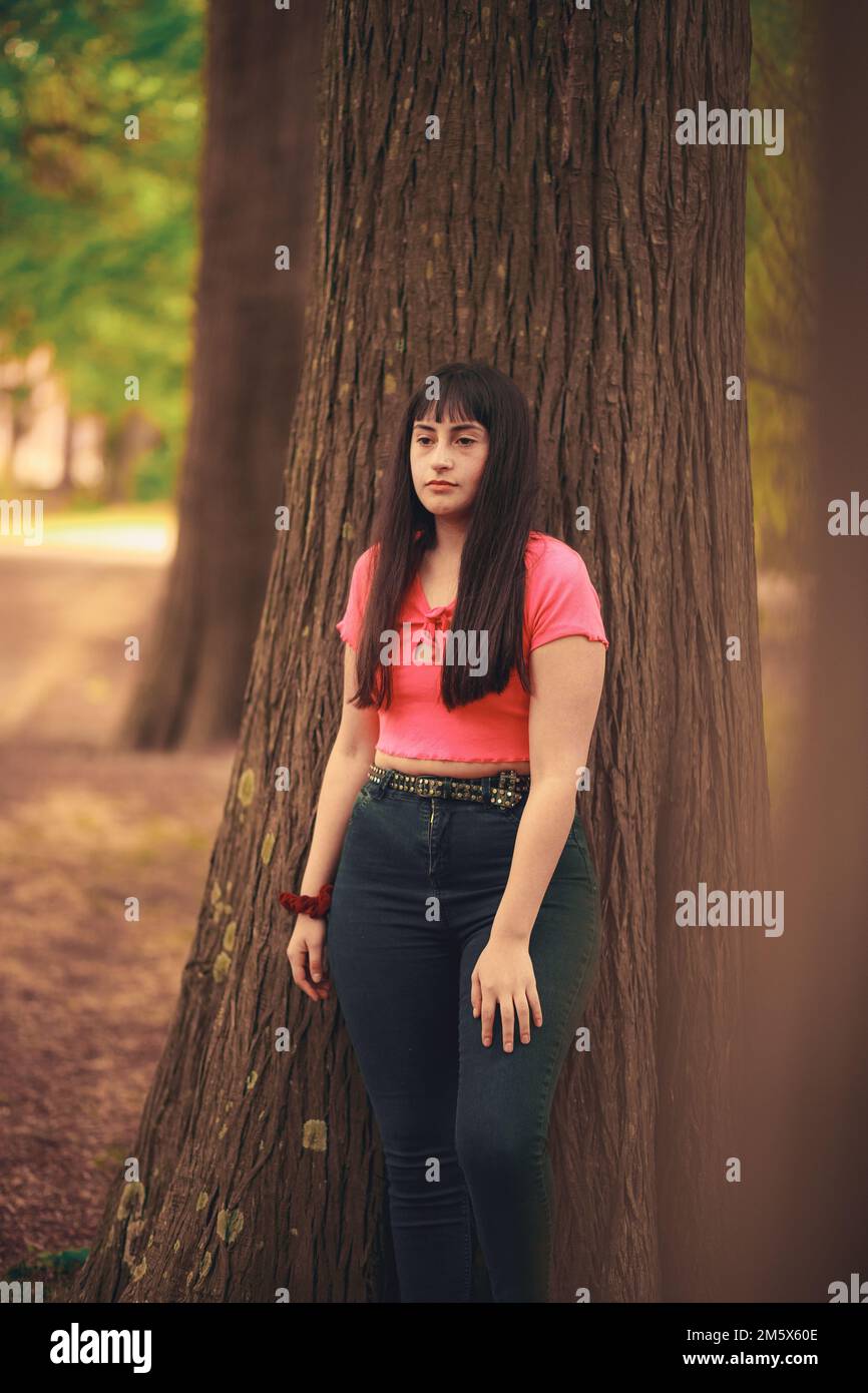 A vertical shot of a beautiful young woman standing behind a tree in a ...
