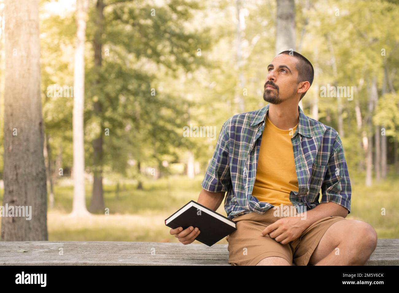 Young man sitting outdoors holding book in a park with trees background ...
