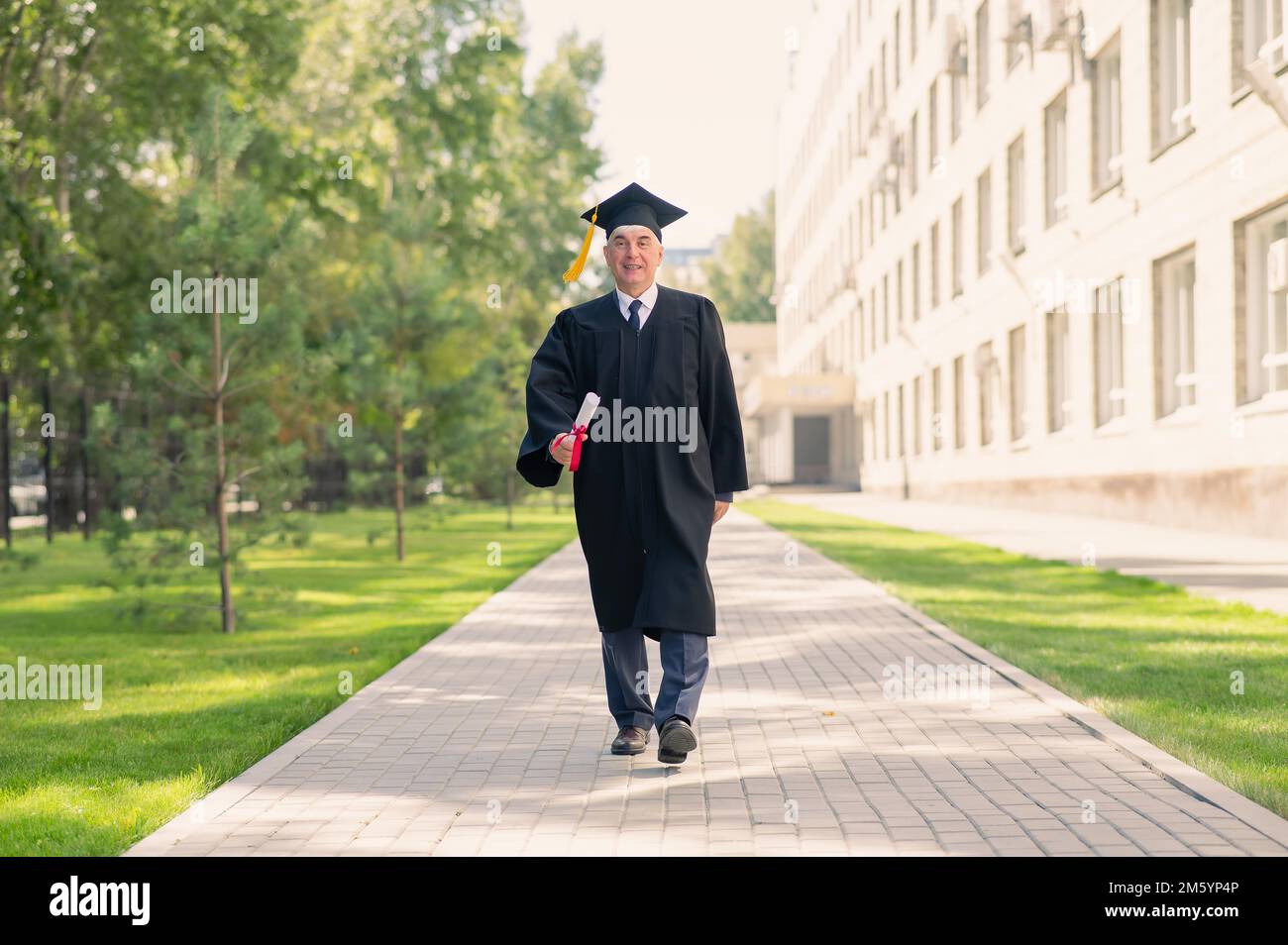 An old man in a graduation gown walks outdoors and holds a diploma ...