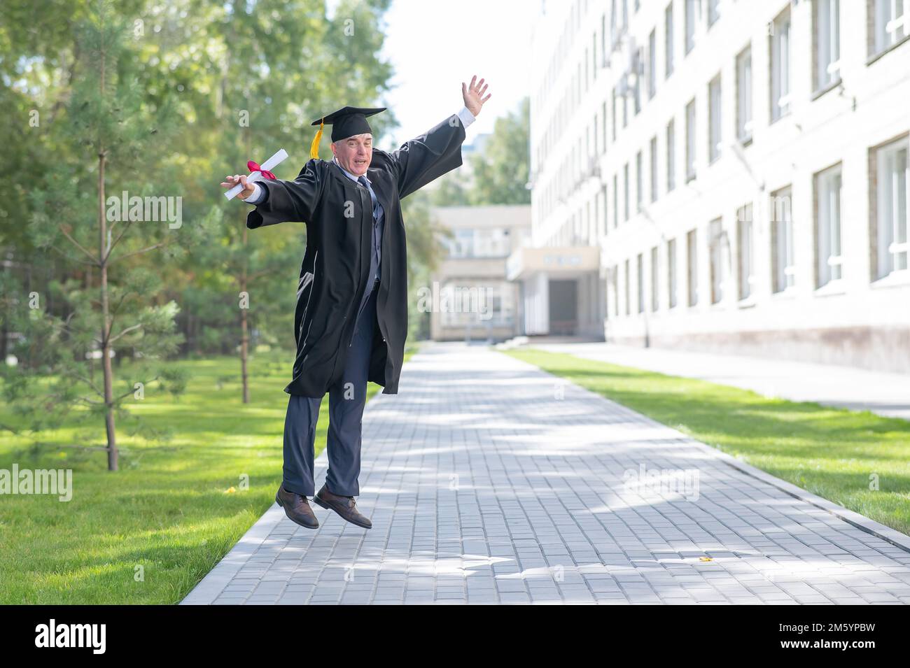 Old happy man in graduation gown jumping outdoors and holding diploma ...