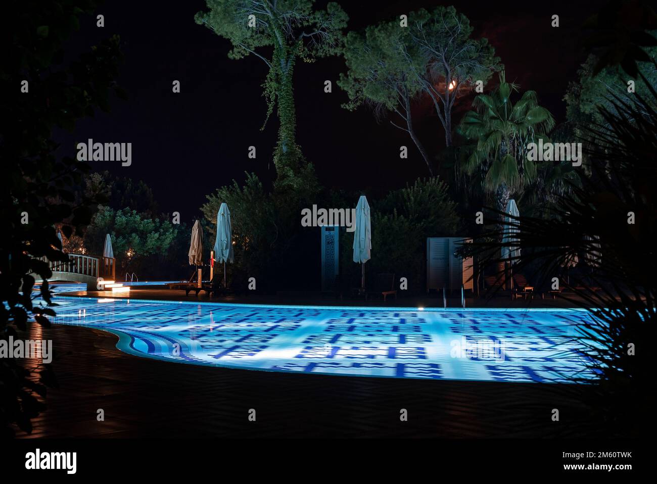 Illuminated swimming pool surrounded with parasols and trees in tourist ...