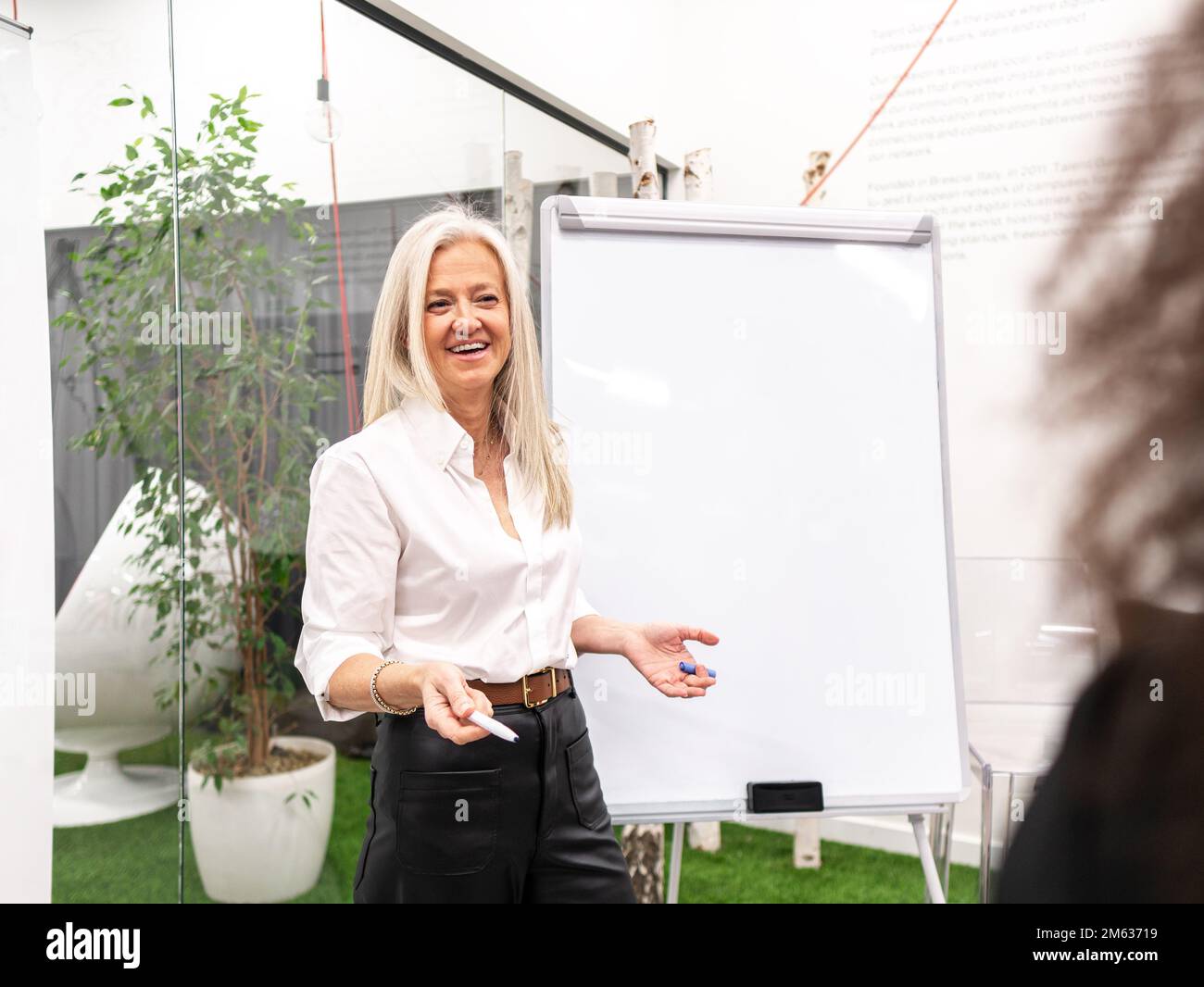 Cheerful senior female boss in formal outfit standing near flip chart ...