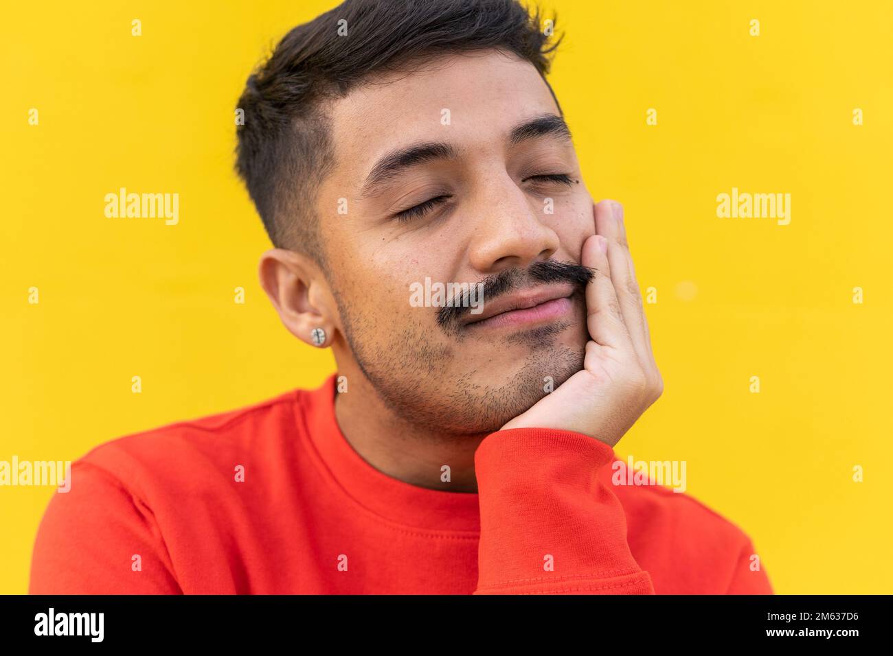 Calm young Hispanic man in red sweatshirt touching cheeks and closing ...