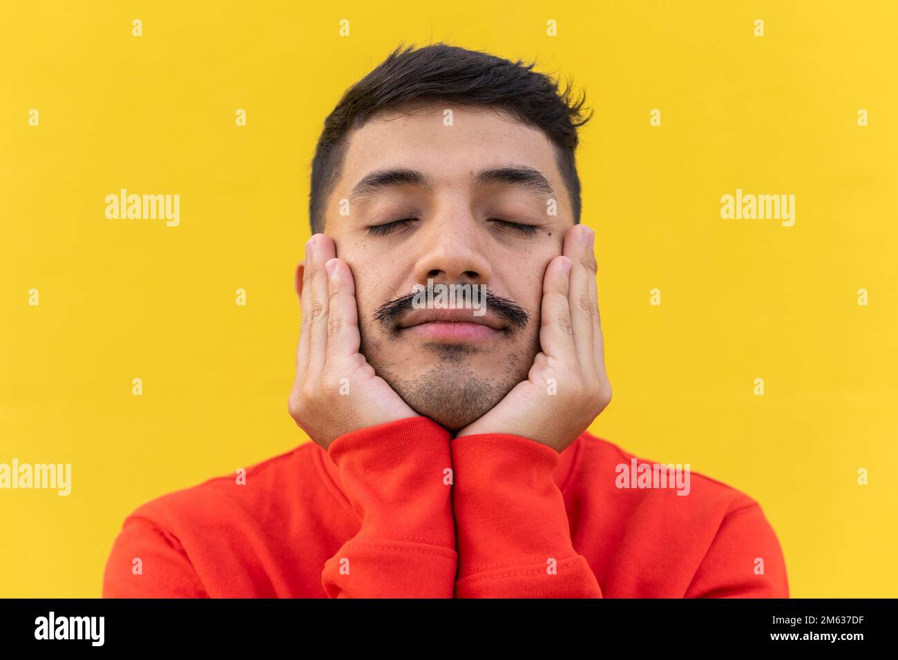 Calm young Hispanic man in red sweatshirt touching cheeks and closing ...