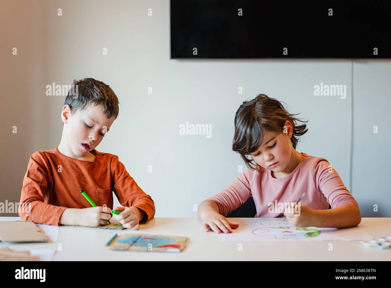 Focused brother and sister in casual clothes sitting at table and creating environmental banners ...