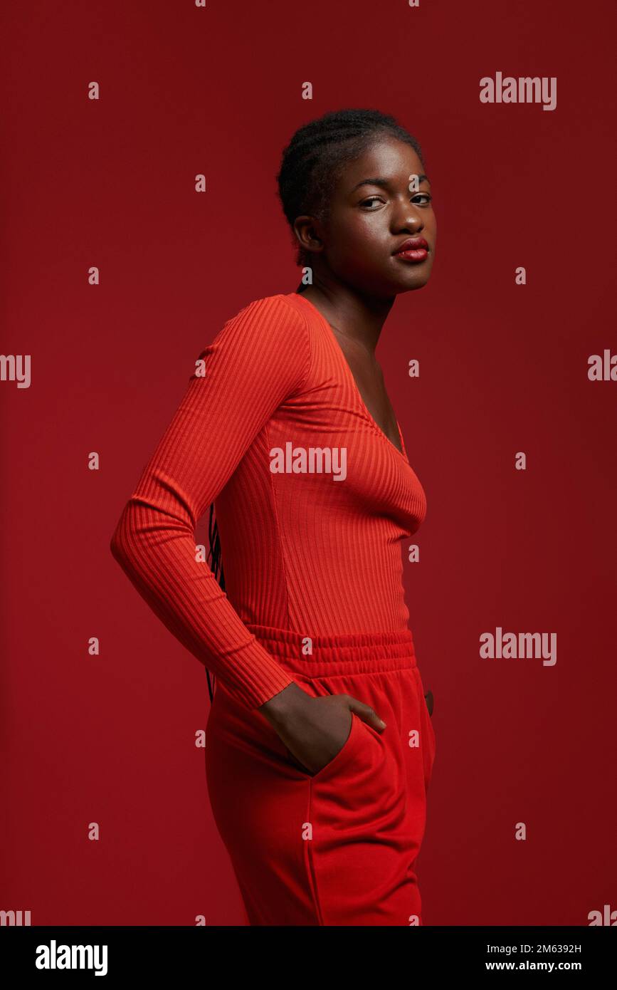 Self assured black female model in dreadlocks wearing total red outfit standing near wall and ...