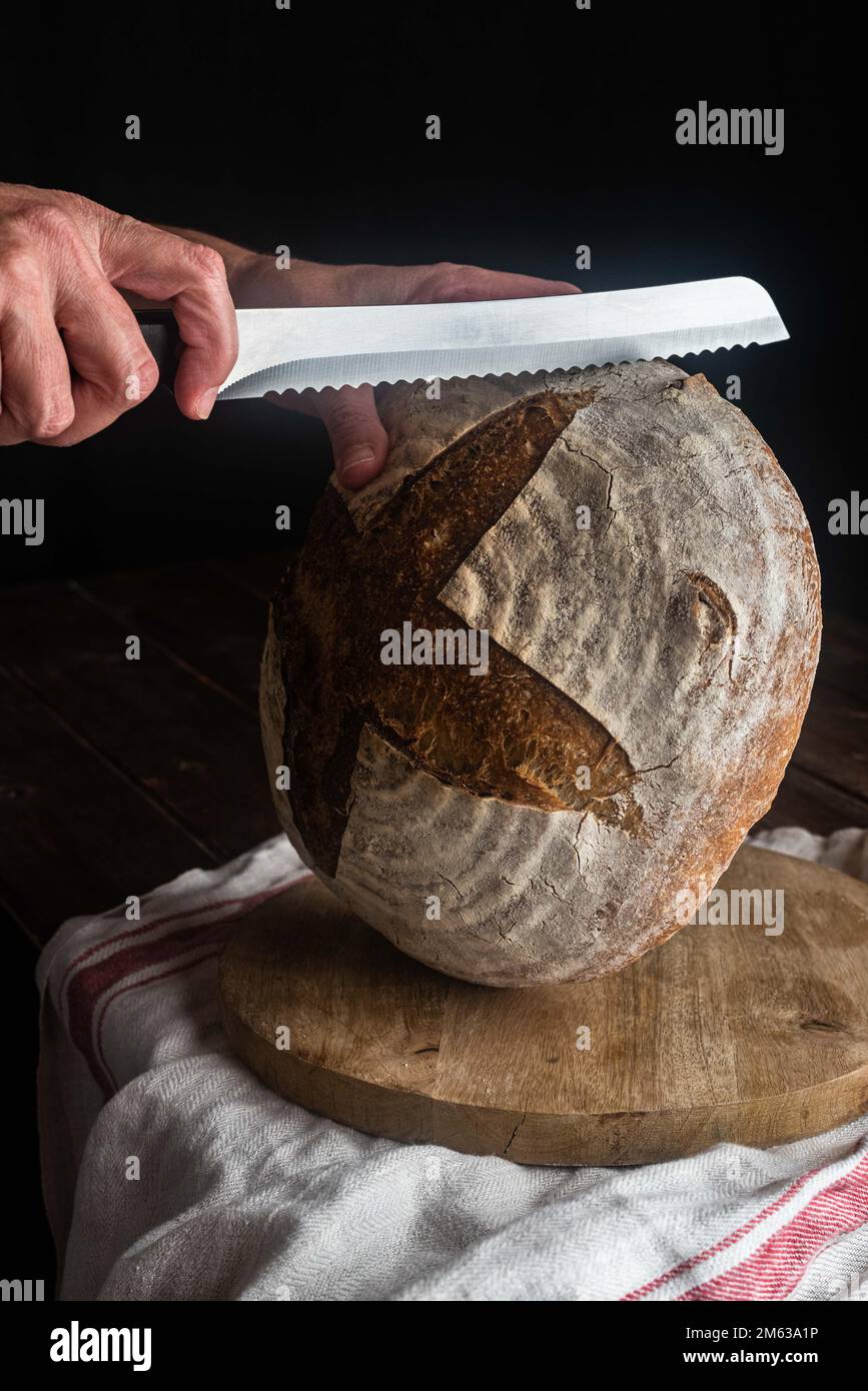 Side view of faceless cook with bread knife cutting whole rye sourdough ...
