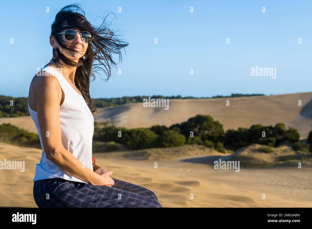 Female with dark hair in sunglasses and casual clothes sitting on sand ...