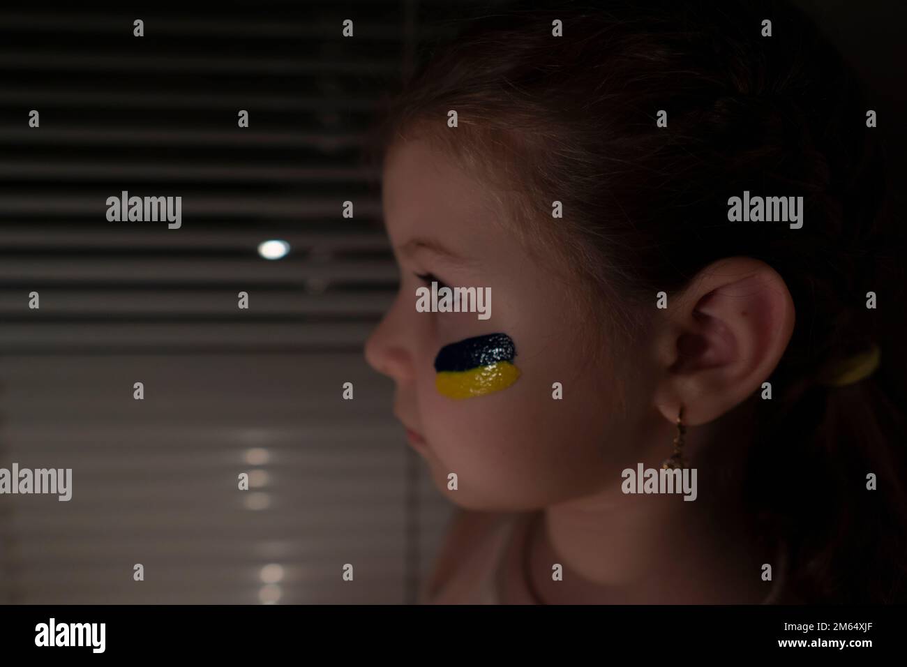 A girl with the flag of Ukraine painted on her cheek stands in a dark ...