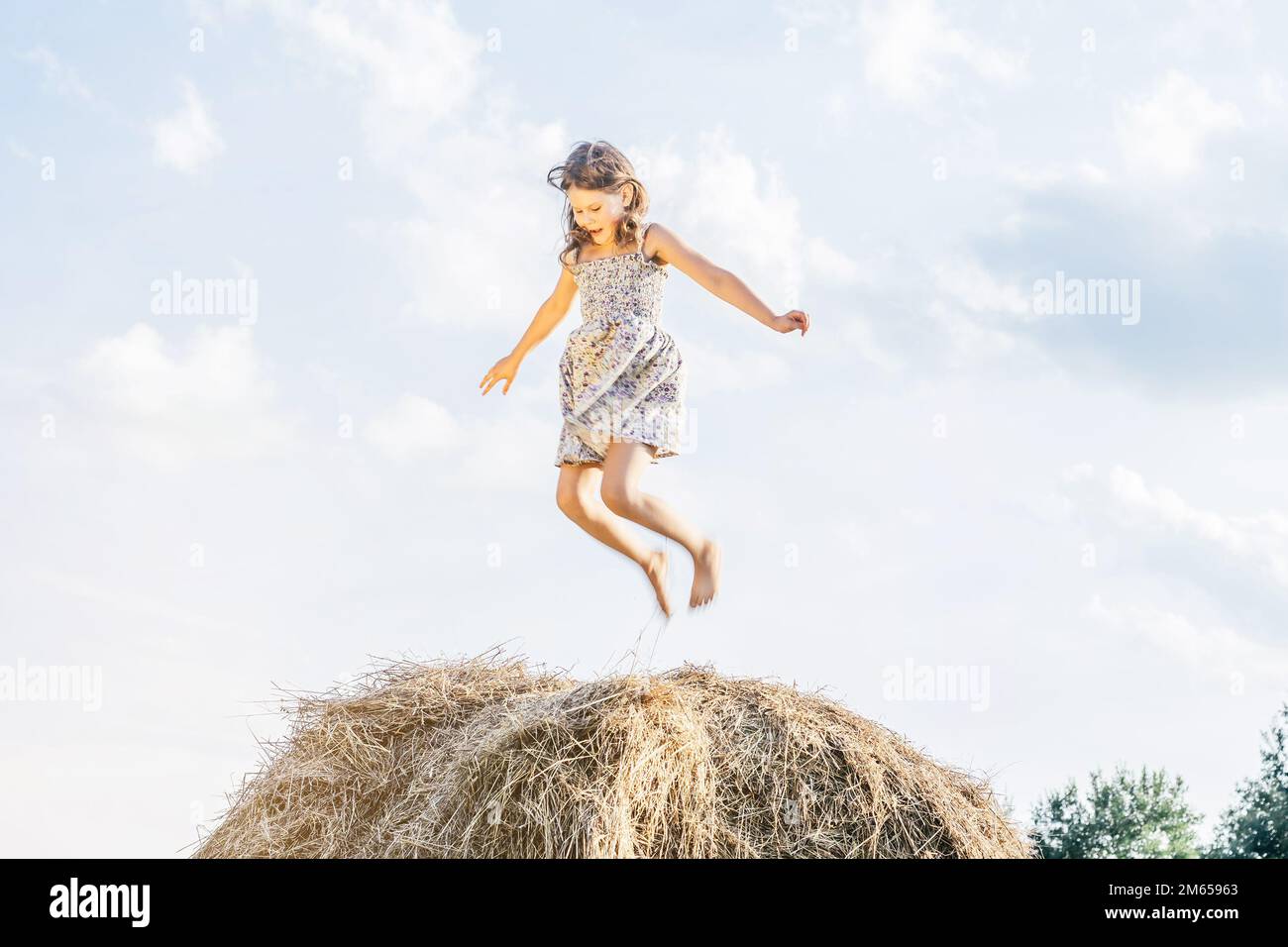 Happy little girl jump up at top of haystack with hands to sides and ...