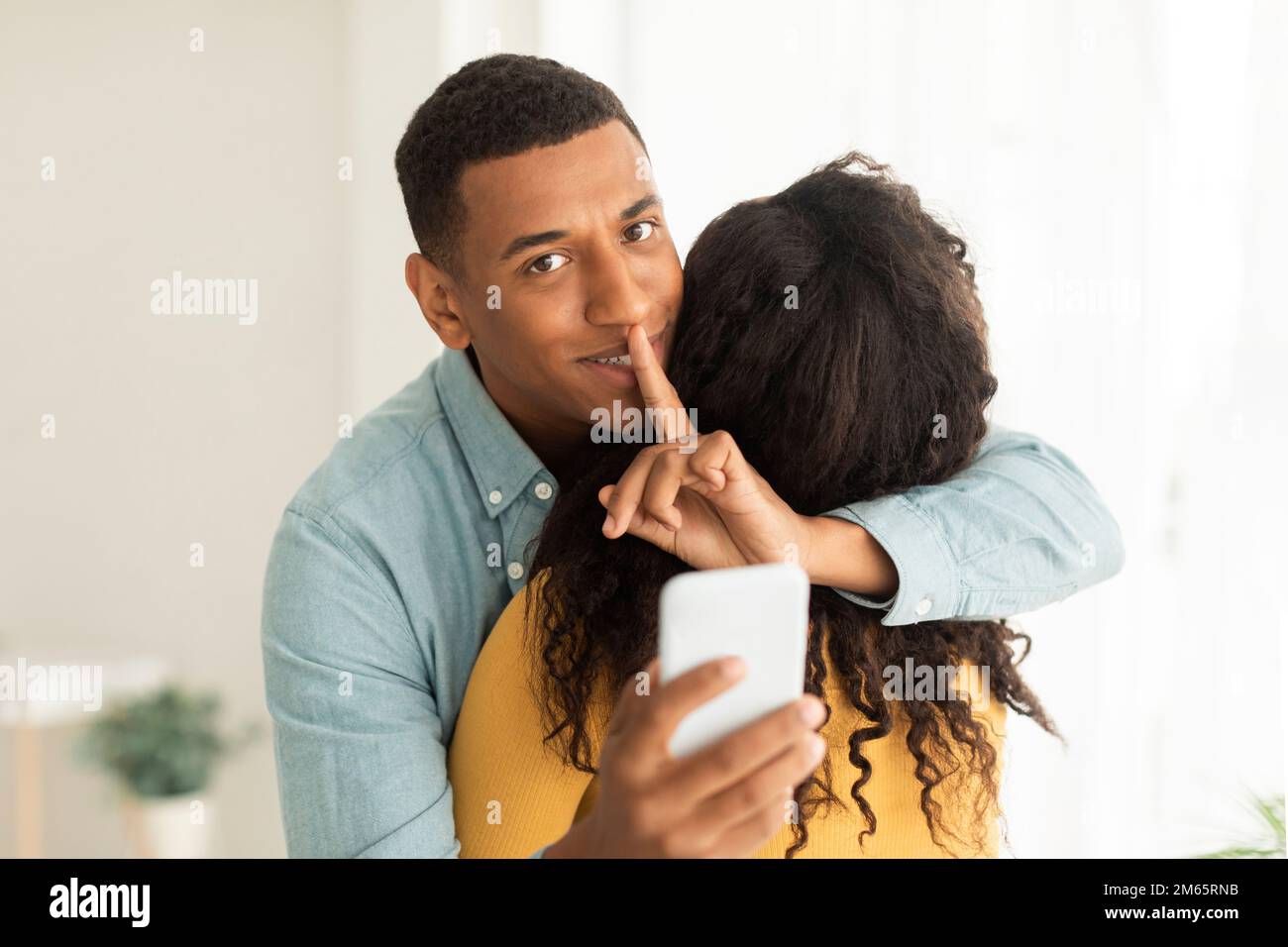Cheerful millennial african american husband hug wife show silence sign, hold smartphone in ...