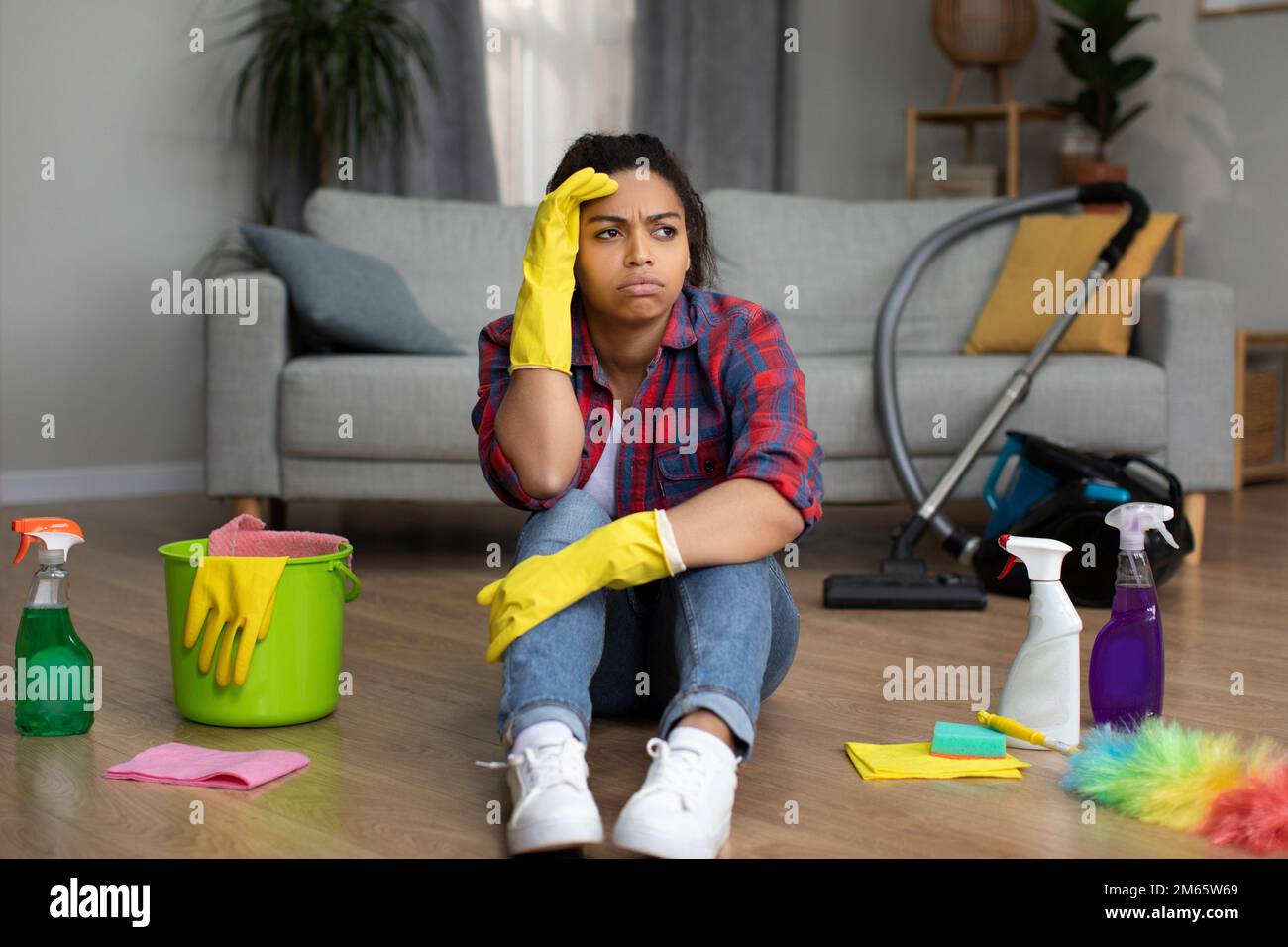 Unhappy tired millennial black lady sit on floor with cleaning supplies ...