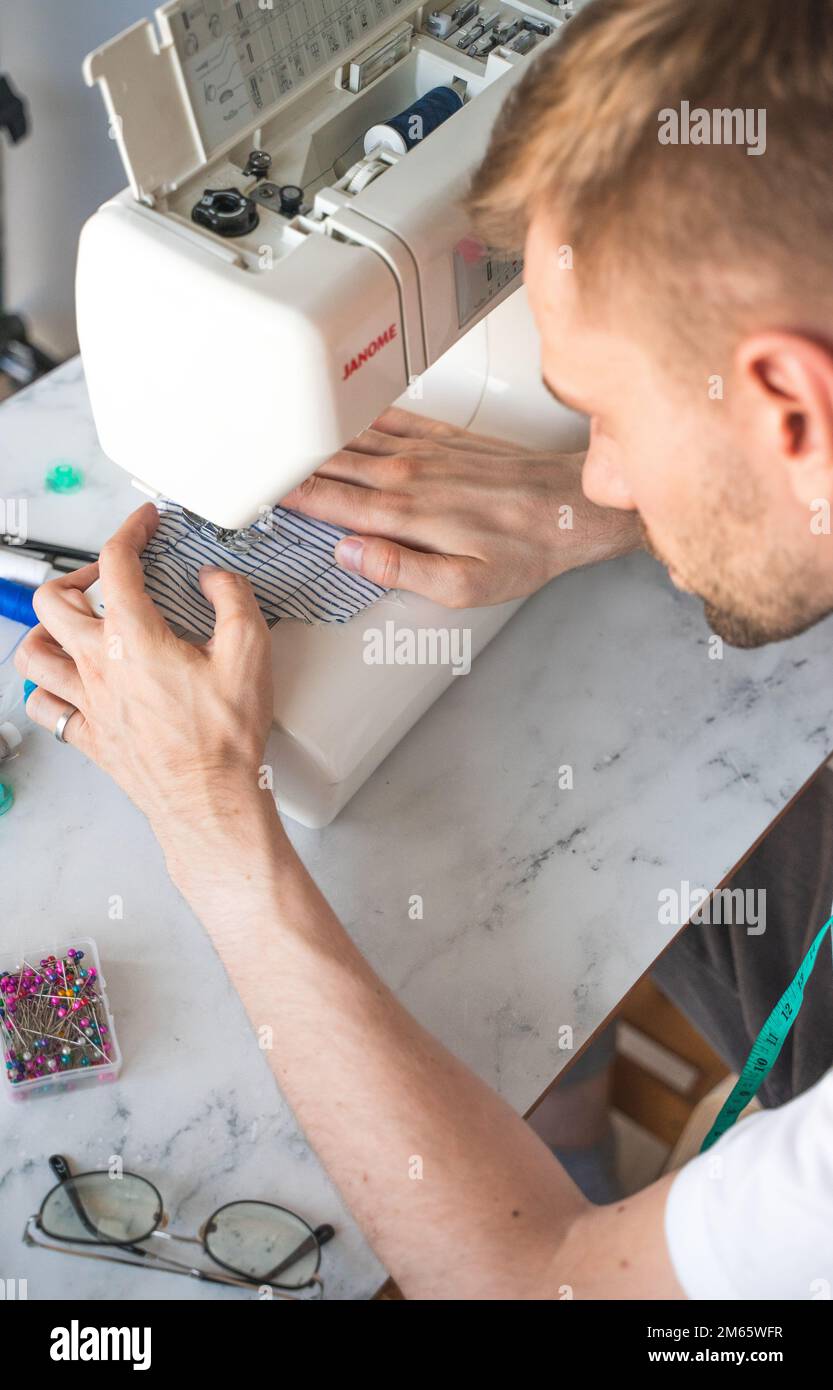 A blond man in white T-shirt is sewing in home workshop. A seamster ...