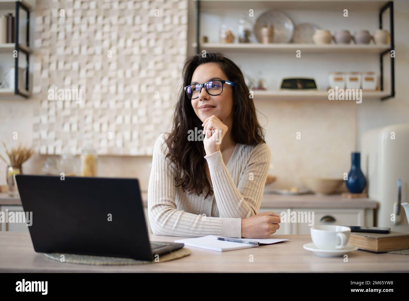 Dreamy woman sitting in kitchen at table with opened laptop, looking ...
