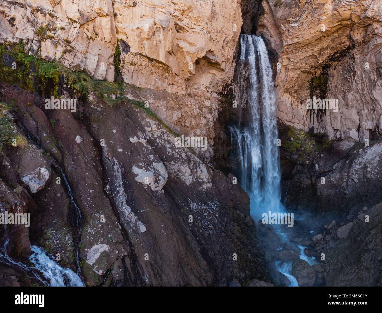 A smooth stream of water flows down from a cave in the high rocks of ...