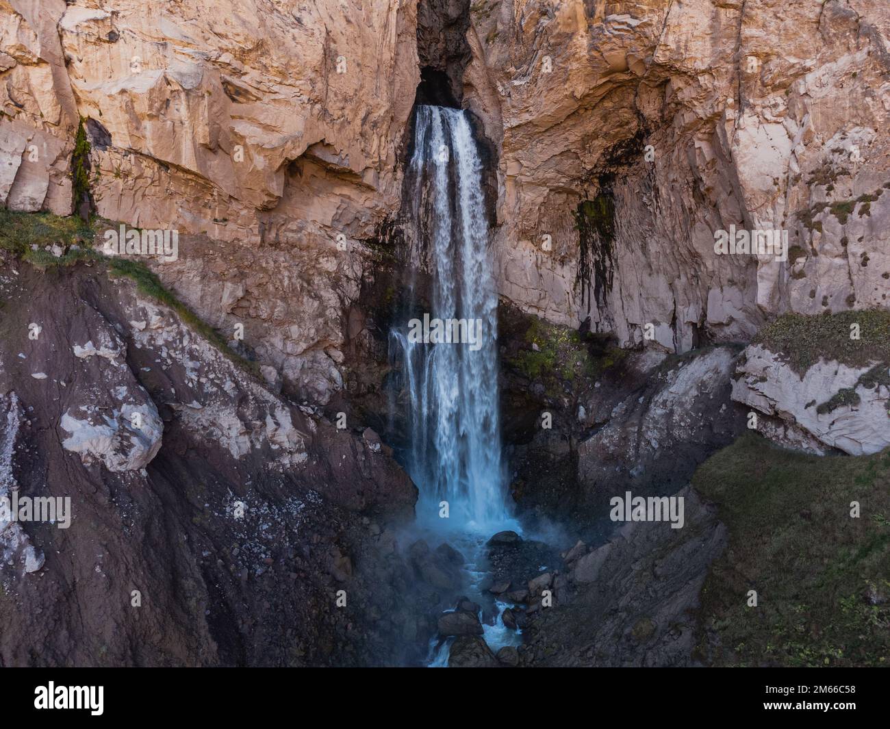 A smooth stream of water flows down from a cave in the high rocks of ...