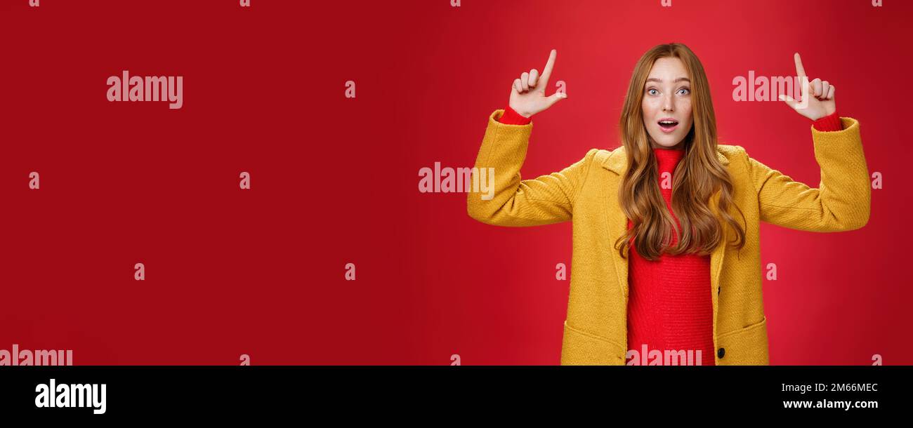 Surprised redhead girl in yellow coat open mouth impressed and thrilled as raising hands and ...